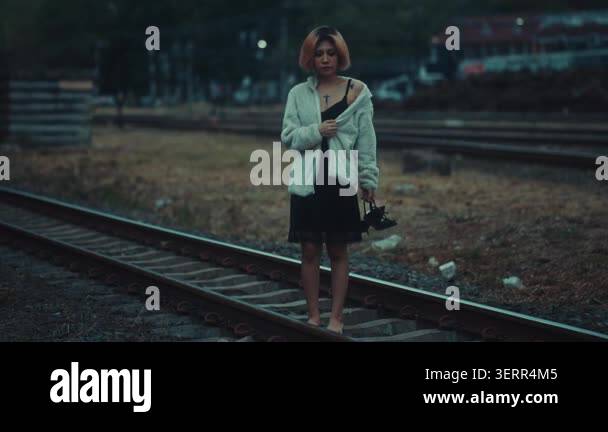 A young woman stands barefoot on gravel railroad tracks in an overcast ...