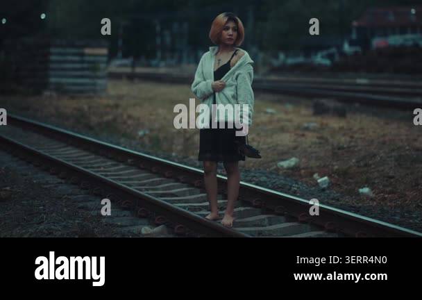 A young woman stands barefoot on gravel railroad tracks in an overcast ...