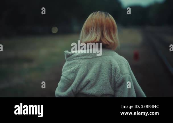 A young blonde woman stands near railway tracks at dusk, slightly ...