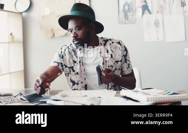 Attractive bearded Afro-American designer in a trendy hat sits by his ...