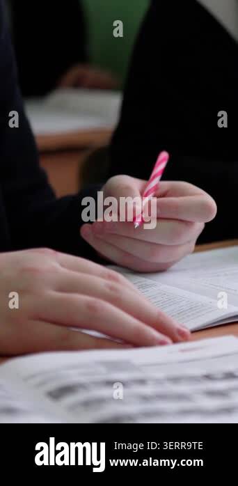 Student holds pink pen while studying textbook on desk, with classmates ...