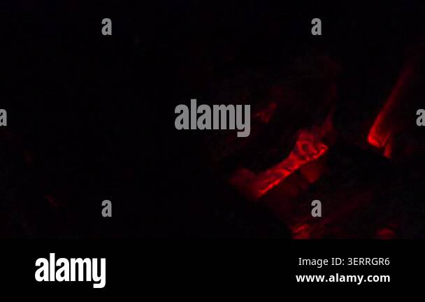 Close-up of glowing red embers and a small flame inside a dark fire pit ...
