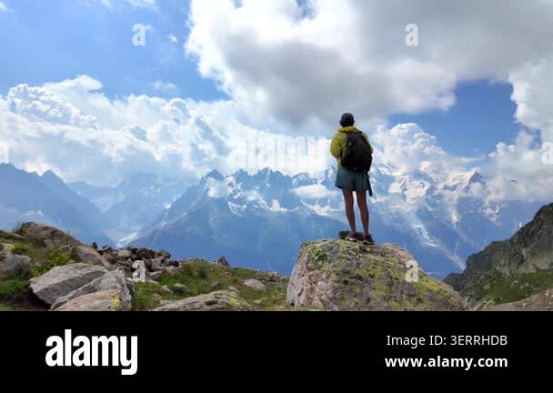 woman hiker stands on a rock, looking out at a vast mountain range ...