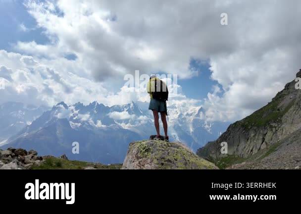 woman hiker stands on a rock, looking out at a vast mountain range ...