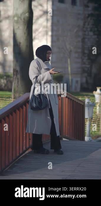 A woman in a coat stands on a bridge looking at her phone in a park ...