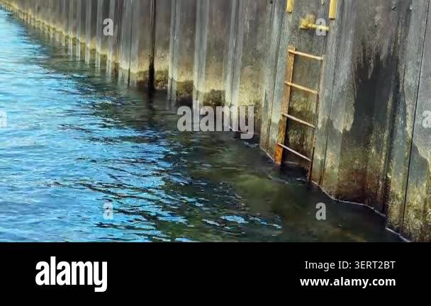Water flows gently against a concrete wall at a port. A ladder stands ...