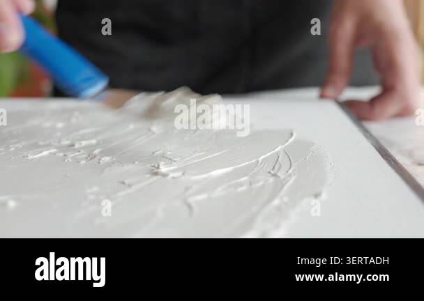 Hand applies white textured paste on canvas with a small spatula ...