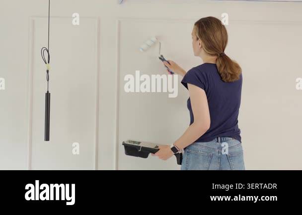 Woman painting wall with paint roller, holding a paint tray while ...