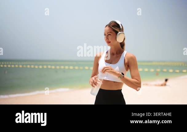 A woman wearing headphones drinks water from a bottle on a beach with ...