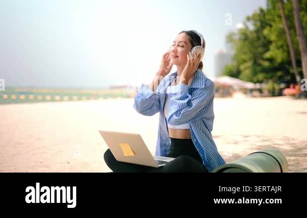 A woman wearing headphones sits on a sandy beach with a laptop ...