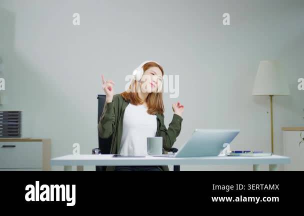 A young woman wearing headphones is sitting at a desk, enjoying music ...