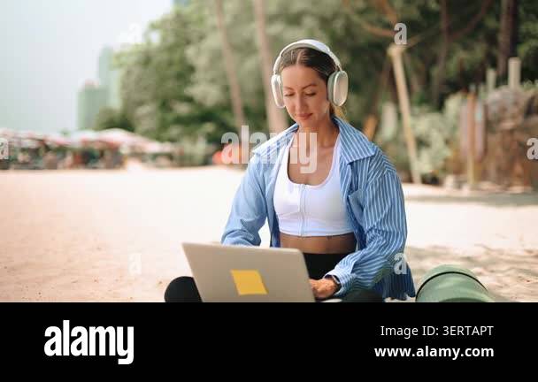A woman wearing headphones is typing on a laptop while sitting on a ...