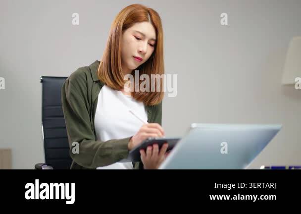 A young woman with auburn hair is sitting at a desk, using a stylus to ...