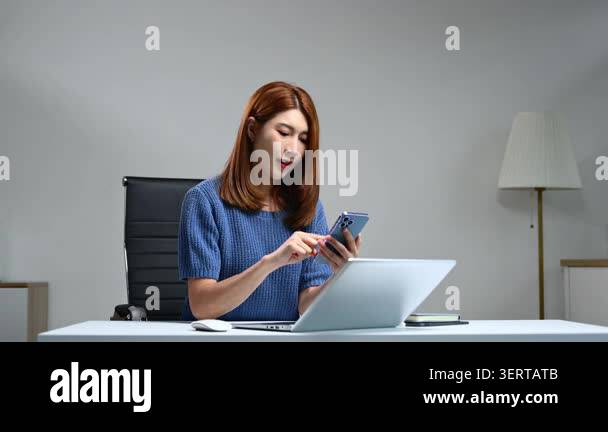 A woman is sitting at a desk, using her smartphone and laptop in a ...