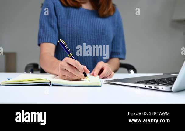 A woman is writing in a notebook with a pen next to a laptop computer ...