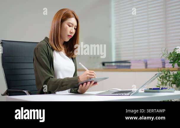 A young woman is sitting at a desk and using a stylus to draw on a ...