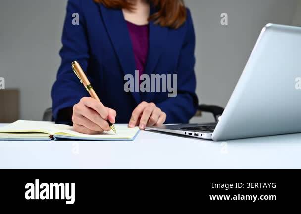 A woman in a blue blazer is writing in a notebook next to a laptop ...