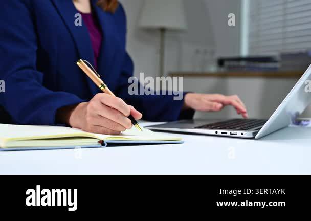 A person in a blue blazer is typing on a laptop and writing in a ...
