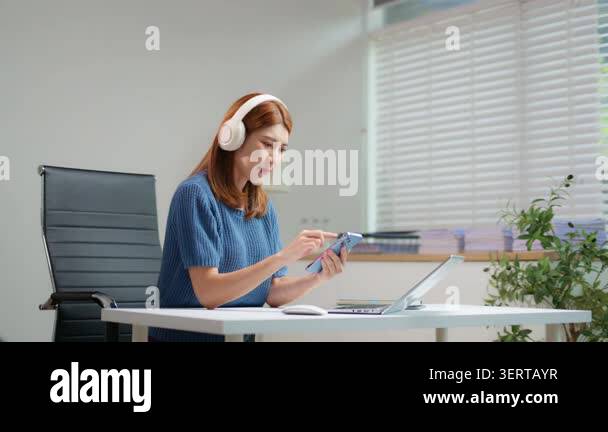 A woman wearing headphones is typing on a laptop while holding a ...
