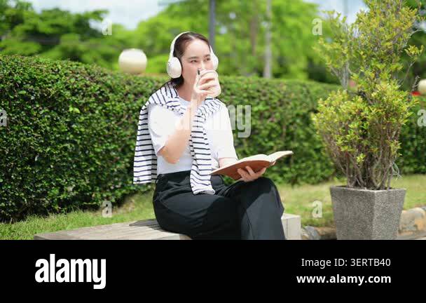 A young woman wearing headphones is sitting on a bench outdoors ...