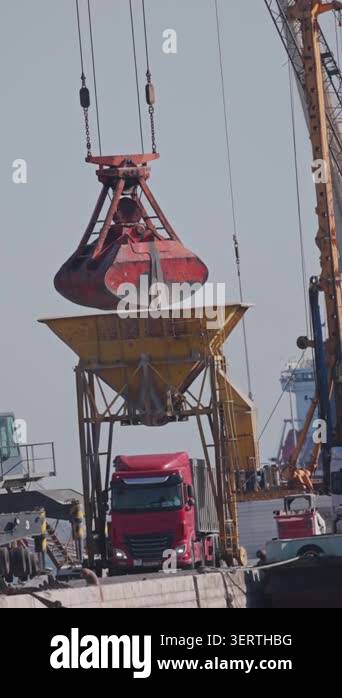 A large crane is using a bucket to lift cargo at a busy shipping port ...