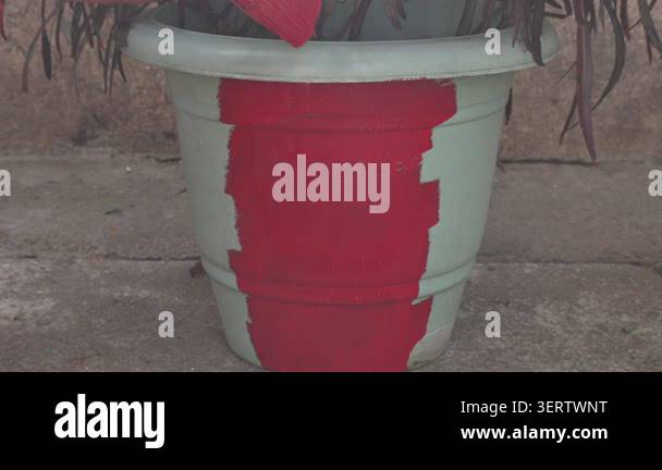 A person uses a paintbrush to apply red paint over a plastic pot. The ...