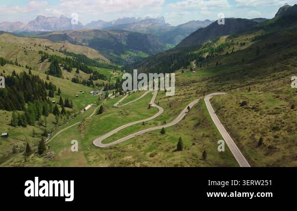 A paved road bends through a wide alpine valley with green meadows ...