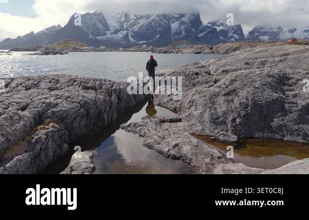A lone hiker in dark outdoor clothing and a red hat stands on craggy ...