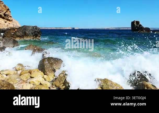 Crystal clear blue ocean waves crash gently against a rocky coastline ...