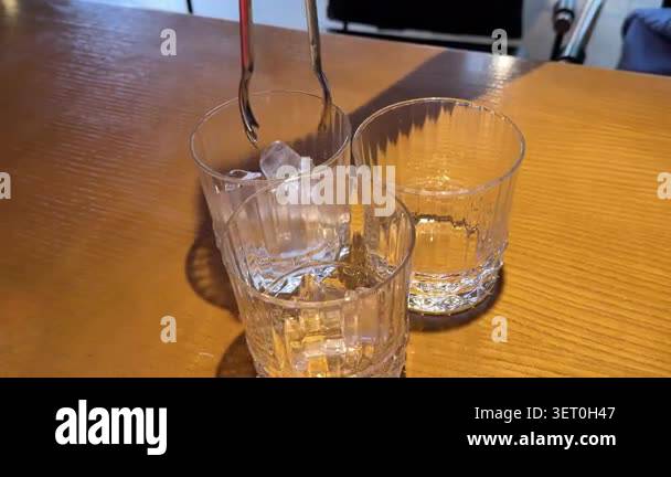 Metal tongs place sparkling ice cubes into empty tumbler preparing a ...