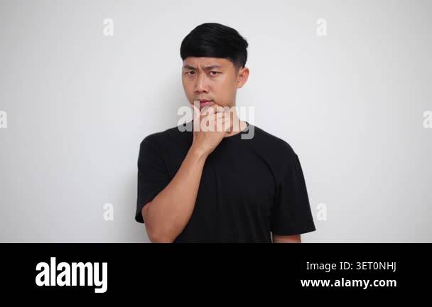 Asian man in a black shirt touches his chin and looks around ...