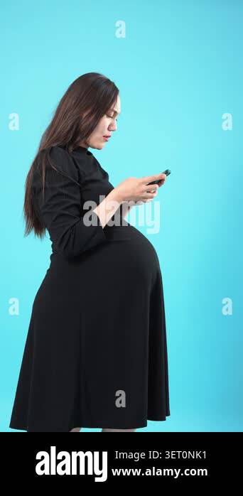 A pregnant female stands sideways using her phone and rubs her head ...