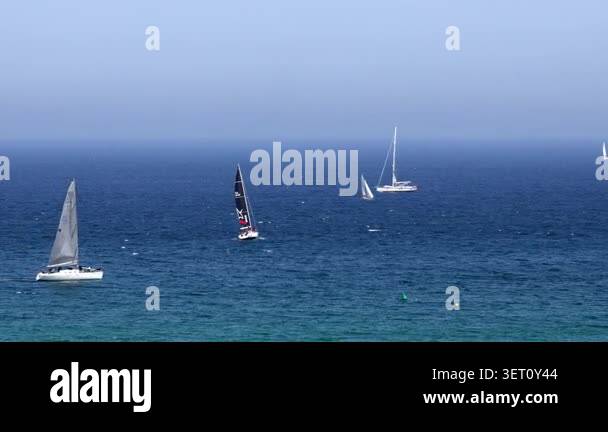 Several sailboats glide across the deep blue ocean under a hazy sky ...