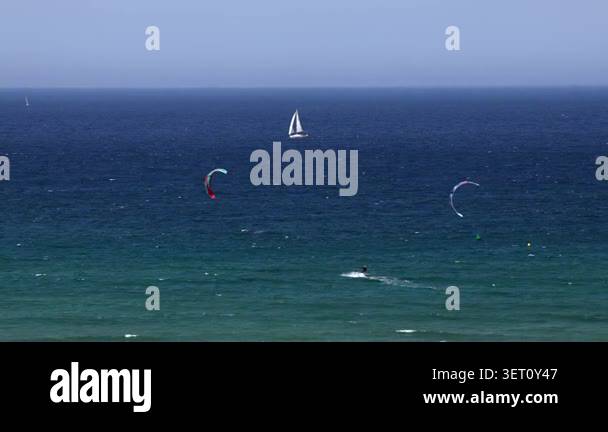 Wide shot of a sailboat and kite surfers on a choppy blue ocean under a ...