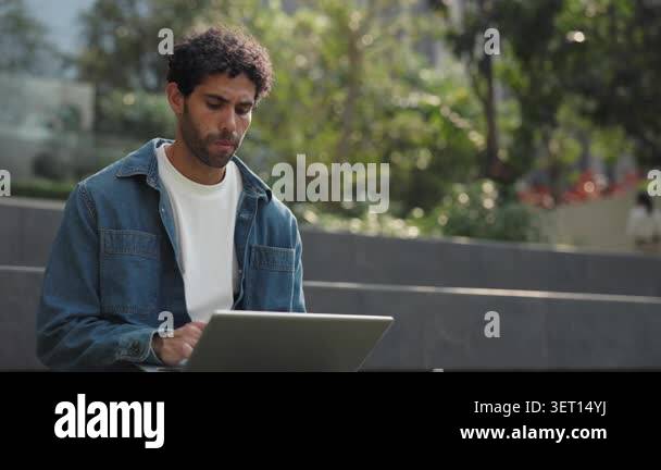 Portrait of focused Hispanic man works on laptop as sits on parapet in ...