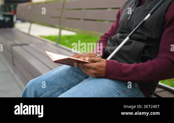 Black man with blindness reading a braille book on a park bench. A ...