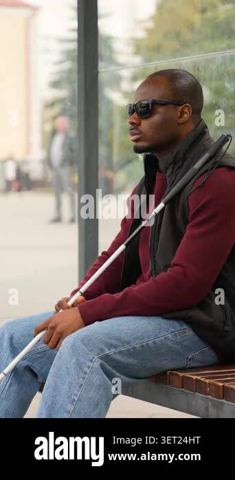 Young black man with a visual impairment sitting on a bench at a public ...