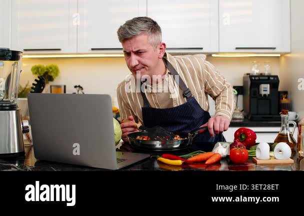 Middle-aged man fries vegetables in pan while watching laptop for diet ...