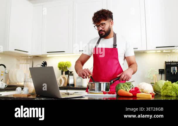 Arabian man stirring soup in pot on stove watching laptop video recipe ...