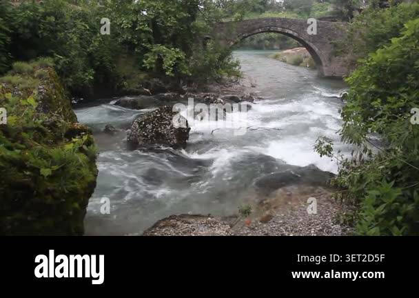 Fast flowing river water rushes over rocks towards an old stone arched ...