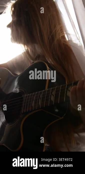 A girl teenager plays an acoustic guitar while sitting by a window. The ...