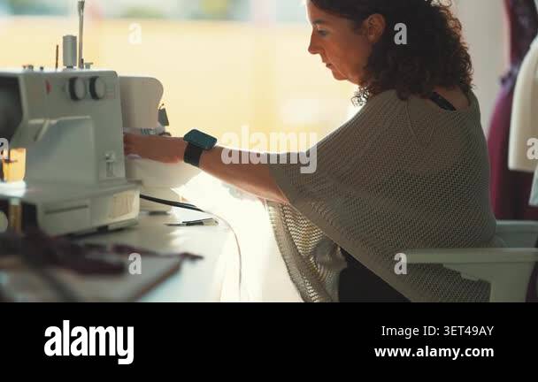 A woman sits at a sewing machine in a well-lit studio. She focuses on ...