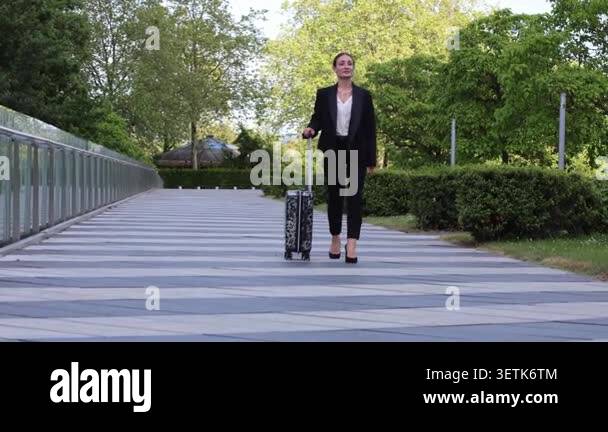 A woman walks along a path in a park. She pulls a suitcase behind her ...