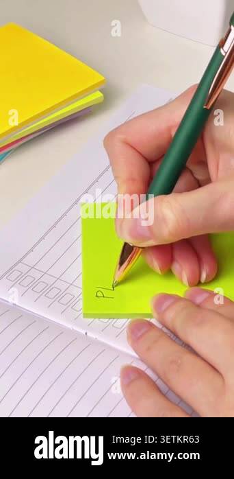 A close-up of a woman's hands writing tasks on a green sticky note ...
