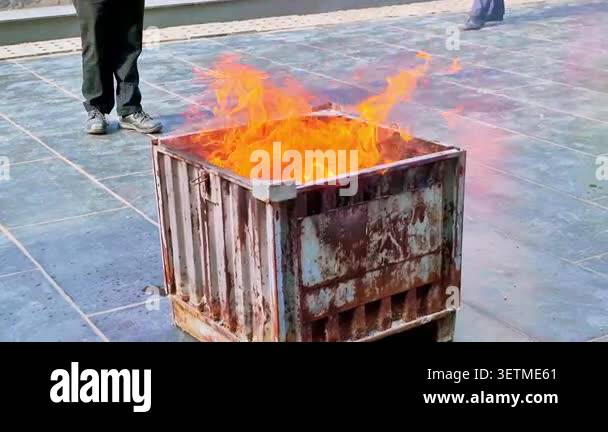A rusted metal burn bin billows thick white smoke as a hose nozzle ...