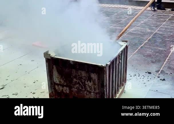 A rusted metal burn bin emits a thick cloud of white smoke as a long ...