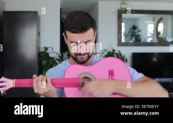 A man is playing a pink guitar indoors, smiling and looking off-camera ...