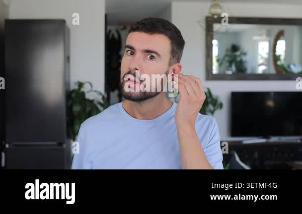 A man is cleaning his ear with a cotton swab in a well-lit room. He is ...