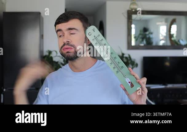 Man holding a thermometer, fanning himself and looking uncomfortable ...