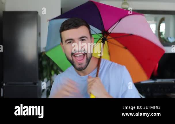 A man with a rainbow umbrella is smiling and dancing indoors ...